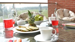 Uma mesa com um jarro de vidro, uma tigela de frutas e um prato com frutas fatiadas, com vista para uma parede de tijolos e vegetação.