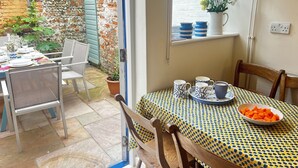 Uma área de refeições com mesa posta para quatro pessoas, cadeiras, um vaso de planta e uma janela com vista para uma parede de tijolos e vegetação.