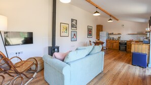 Sala de estar com sofá, televisão e lareira. Há uma área de cozinha com armários de madeira e geladeira.