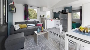 Um apartamento moderno com um sofá cinzento, uma mesa de jantar com frutas, uma cozinha com frigorífico e uma janela ampla.
