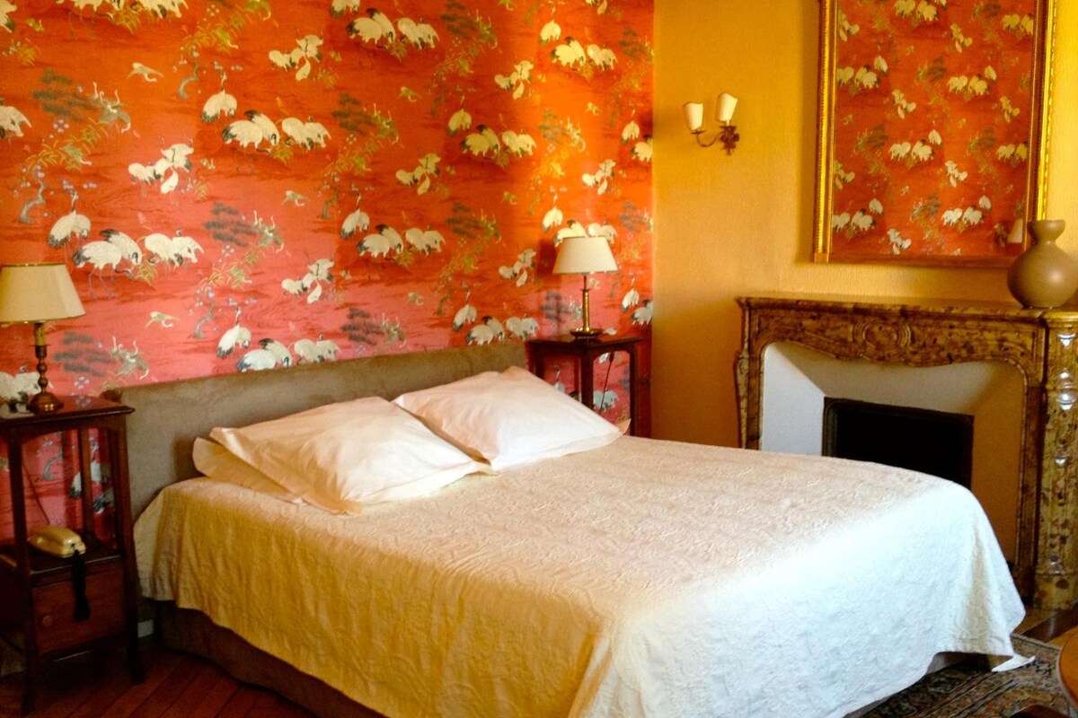 Cama com colcha branca, duas luminárias de cabeceira, mesa de cabeceira de madeira e papel de parede estampado com ovelhas.