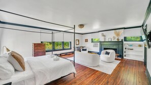 Quarto moderno com cama de dossel, cômoda de madeira, lareira e um espaço de estar com sofá e poltronas brancas.