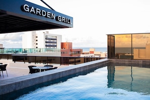 Área de piscina no terraço com um letreiro 'Garden Grill', mesas ao ar livre e vista para o oceano.