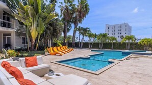 Uma área de piscina com cadeiras de descanso brancas e almofadas laranja, cercada por palmeiras e um edifício ao fundo.