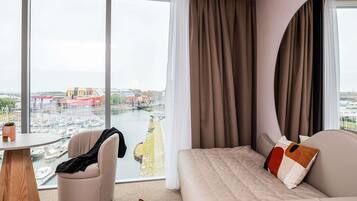 Quarto de hotel com uma janela grande, um sofá, uma cadeira, uma mesa pequena e um par de sapatos no chão.