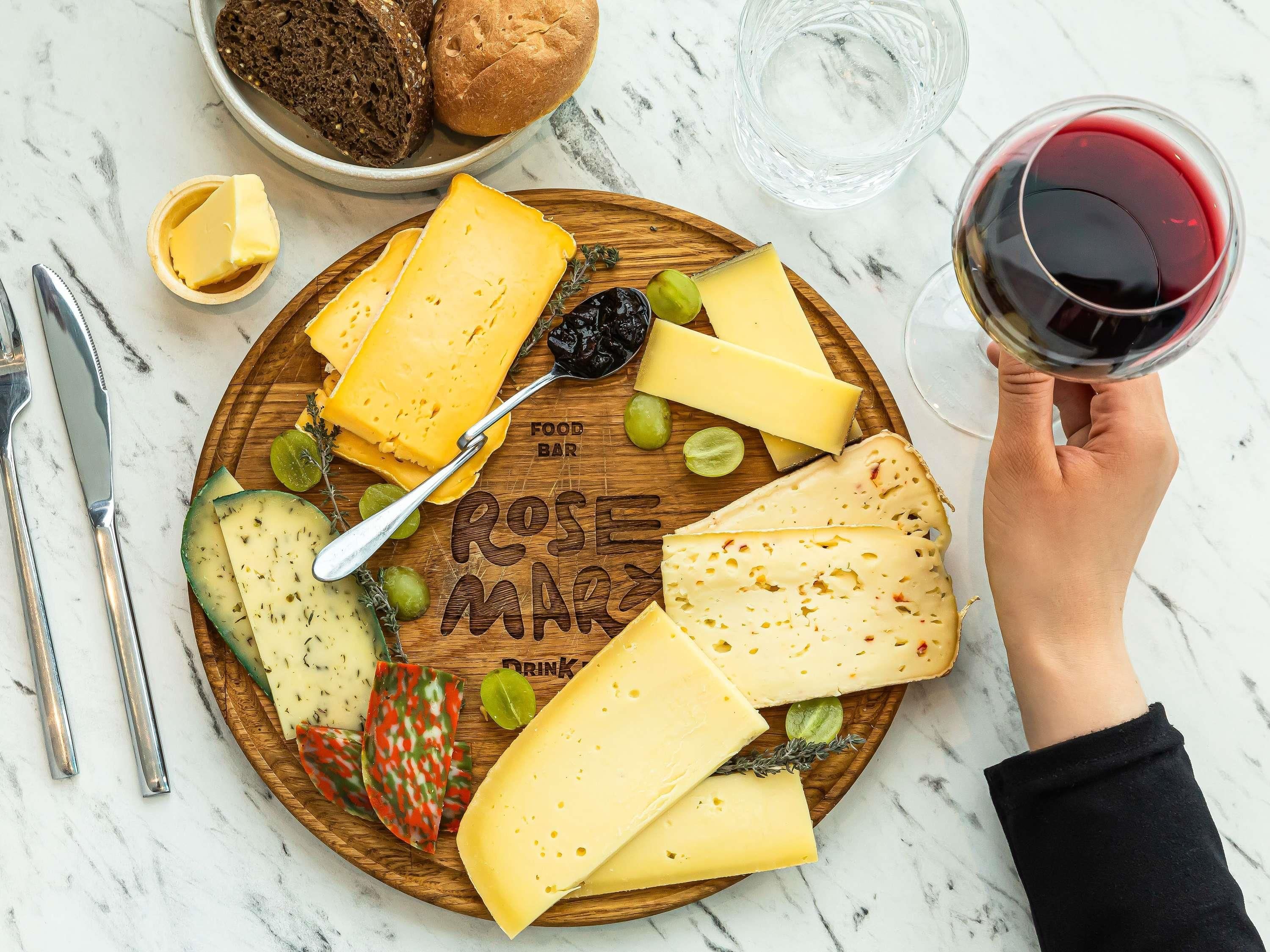 Uma tábua de queijos com diversos tipos, uma garrafa de vinho tinto e uma cesta de pão sobre uma superfície de mármore.