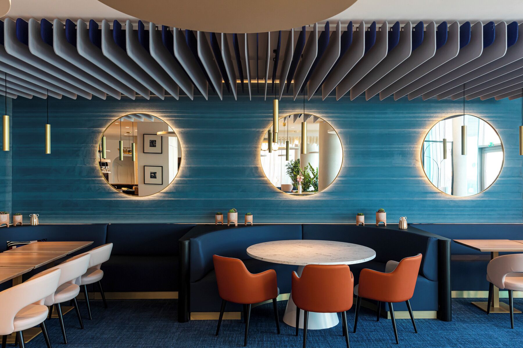 Um restaurante moderno com esquema de cores azul e branco, mesas redondas e cadeiras laranja, e um design de teto único.