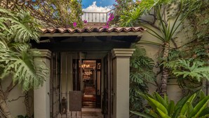 Uma entrada de prédio tradicional com telhado de azulejos, uma placa acolhedora e exuberantes plantas tropicais.