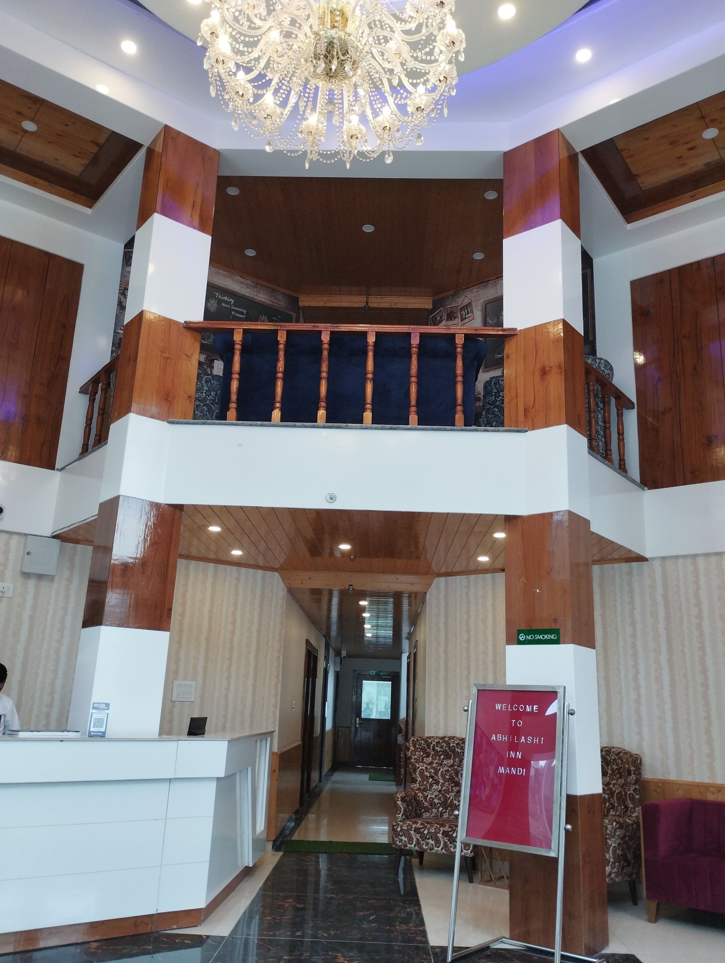 Um hall de hotel com um candelabro, uma mesa de recepção, um aviso de proibido fumar e uma placa de boas-vindas.