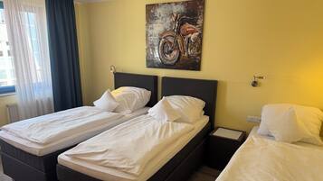 Quarto de hotel com duas camas de solteiro, mesa de cabeceira, quadro na parede e janela com cortinas.
