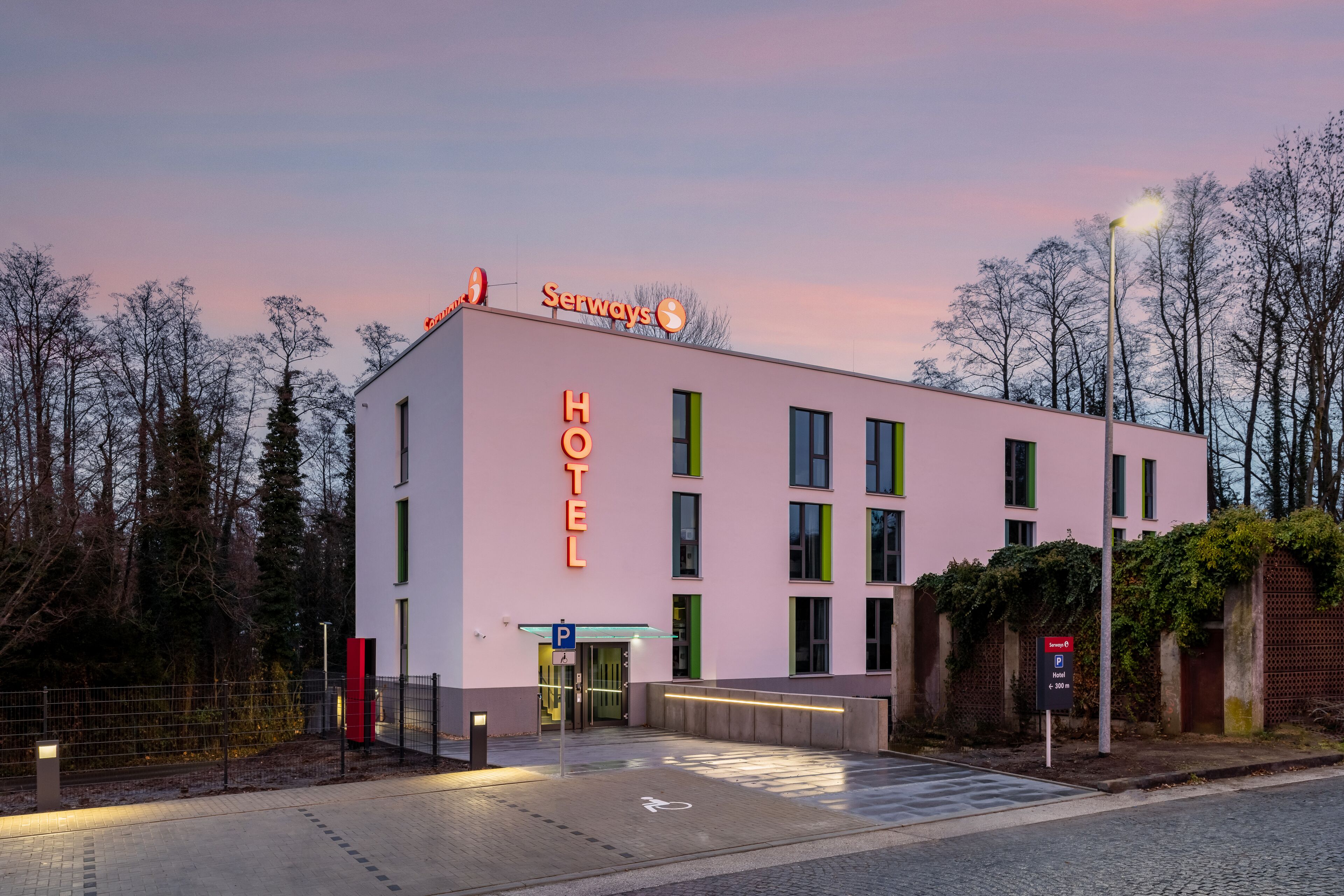 Um hotel moderno com um letreiro chamativo 'HOTEL', uma área de estacionamento e uma placa 'Serways' indicando serviços próximos.