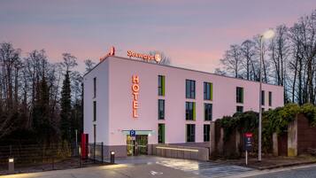 Um hotel moderno com um letreiro chamativo 'HOTEL', uma área de estacionamento e uma placa 'Serways' indicando serviços próximos.