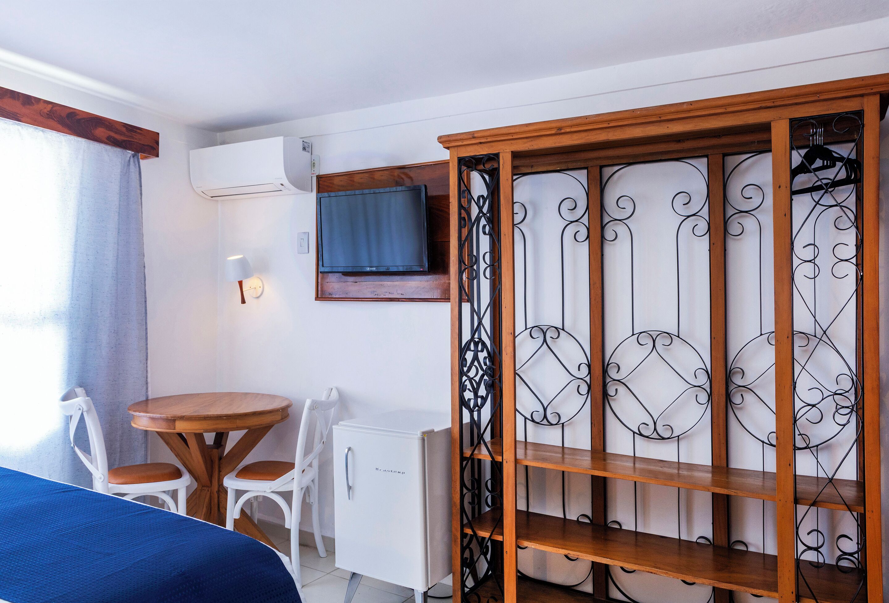 Quarto de hotel com cama, pequena mesa e duas cadeiras, televisão fixada na parede e aparador de madeira com prateleiras.