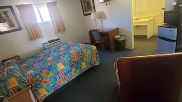 Quarto de hotel com cama, escrivaninha, cadeira, televisão, mini-frigorífico e banheiro visível por uma porta aberta.