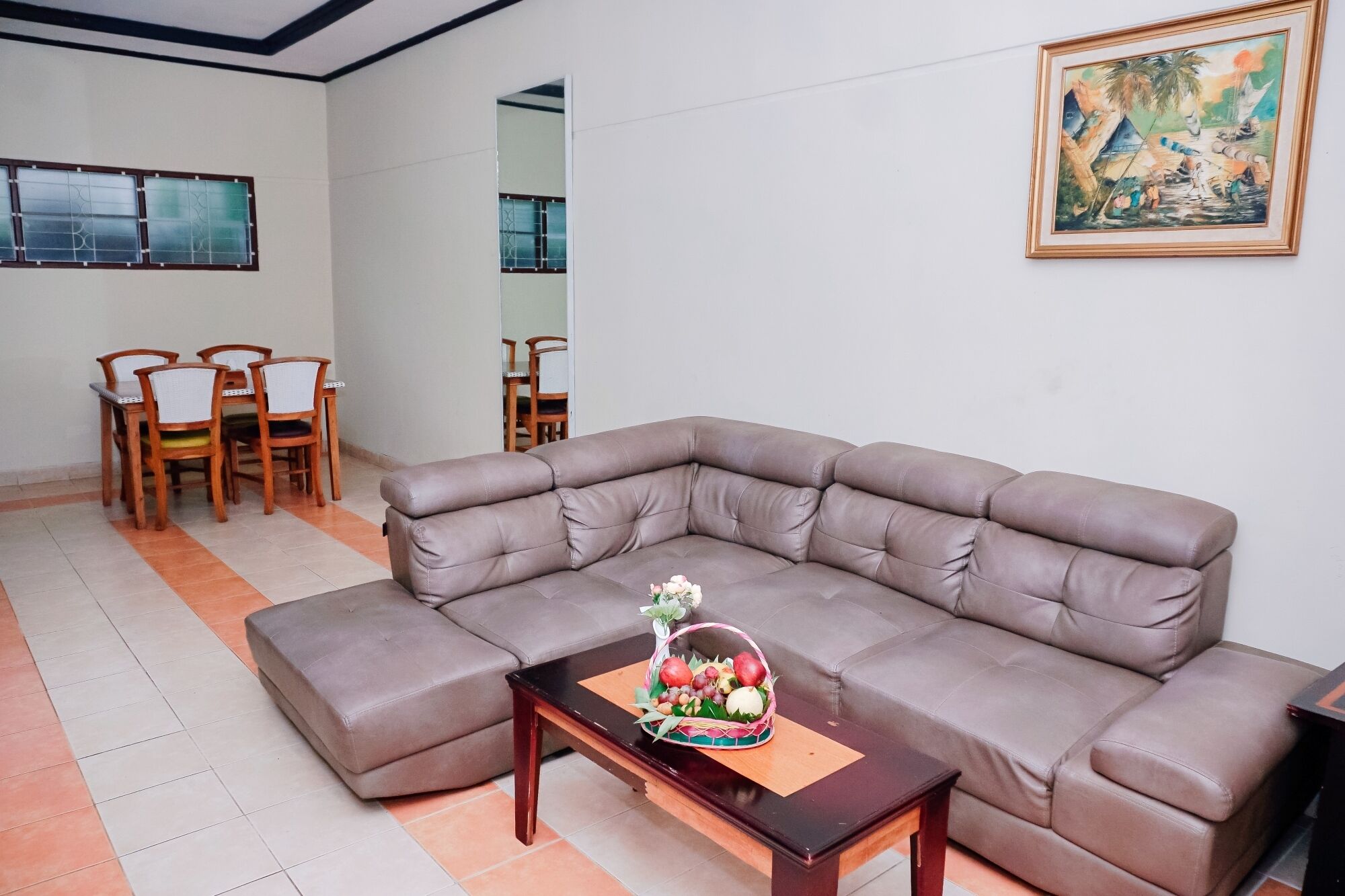 Uma sala de estar com um sofá seccional de couro marrom, uma mesa de centro de madeira com uma cesta de frutas e uma área de refeições com cadeiras e mesa de madeira.