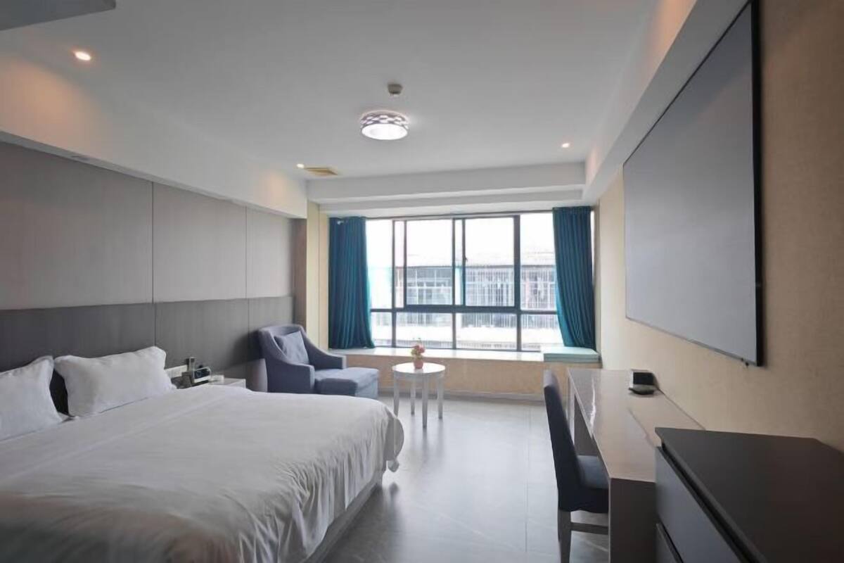 Quarto de hotel moderno com uma cama grande, uma mesa de escritório, uma cadeira, uma mesinha e uma janela ampla com cortinas.