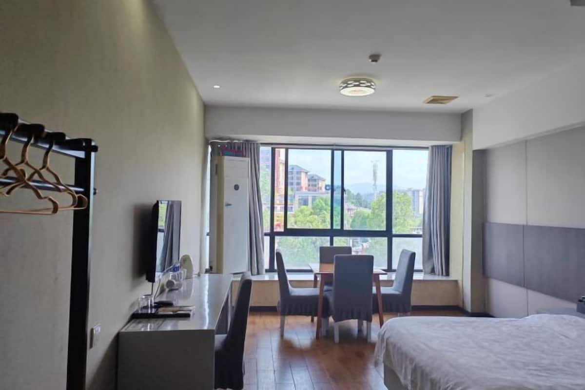 Quarto de hotel com cama, escrivaninha com cadeira, televisão e uma ampla janela com vista para prédios e árvores.