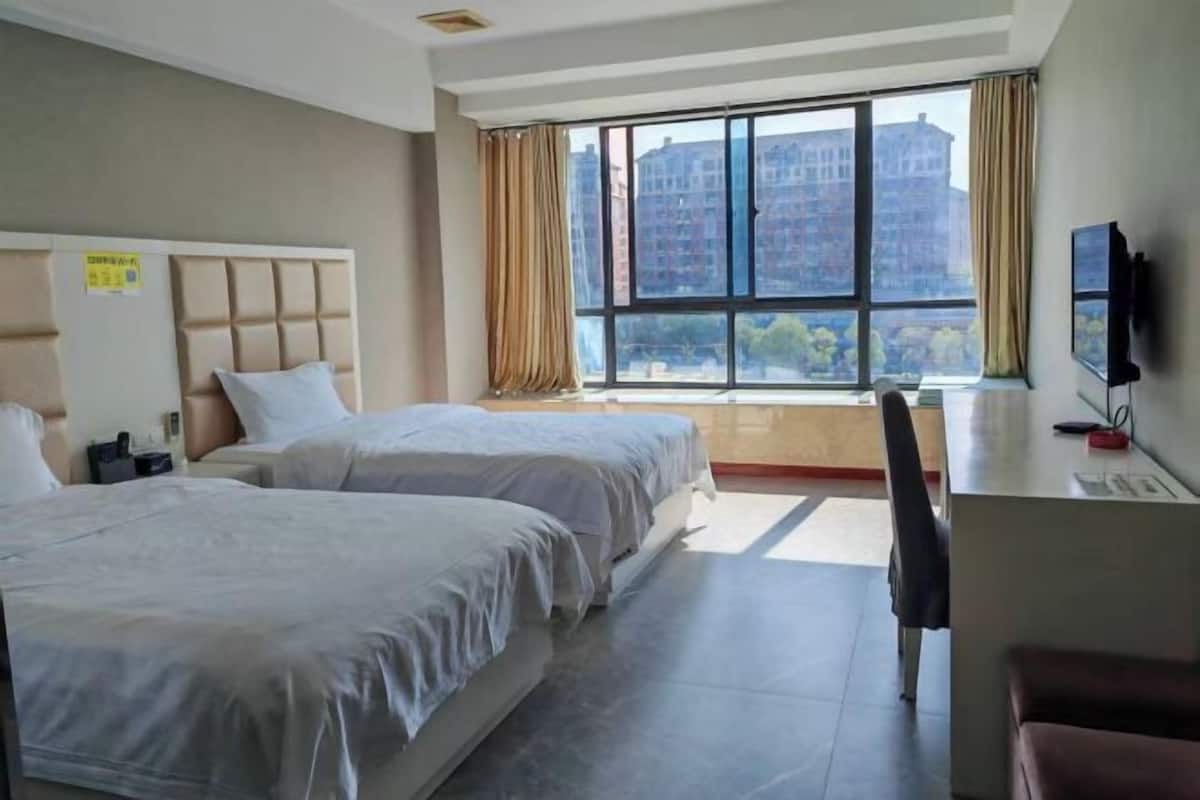 Quarto de hotel com duas camas, uma escrivaninha, uma cadeira, televisão e uma janela grande com vista para a cidade.