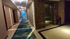 Um corredor de hotel com um pilar de mármore, um elevador de vidro e piso revestido de carpete.