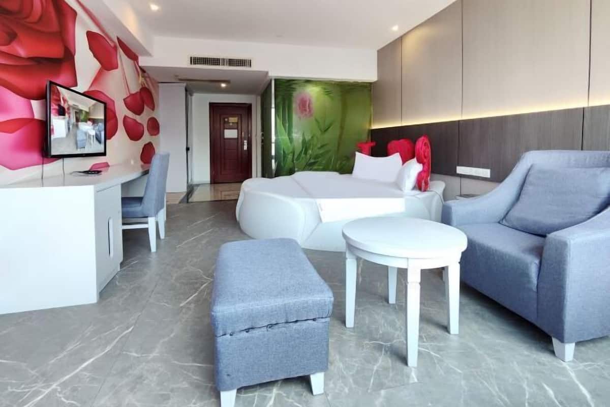 Quarto de hotel moderno com uma cama grande, área de estar com sofá e apoio para os pés, uma mesinha e televisão fixada na parede.