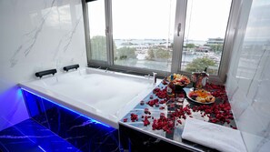 Casa de banho moderna com uma banheira grande, uma janela com vista para a cidade e uma mesa com frutas e flores.