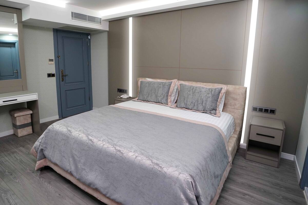 Um quarto com cama, mesas de cabeceira, cômoda, espelho e porta.