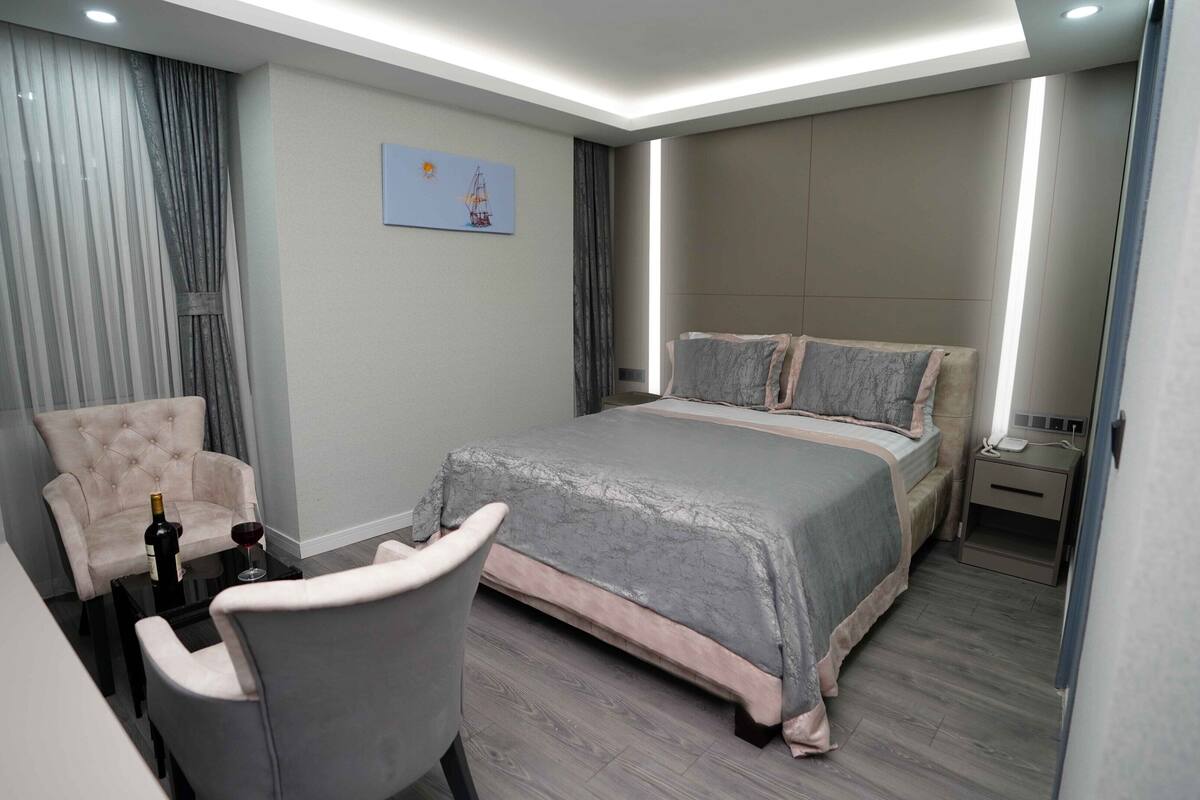 Quarto de hotel moderno com cama, uma cadeira, mesa de cabeceira com garrafa e copo, e um quadro na parede.