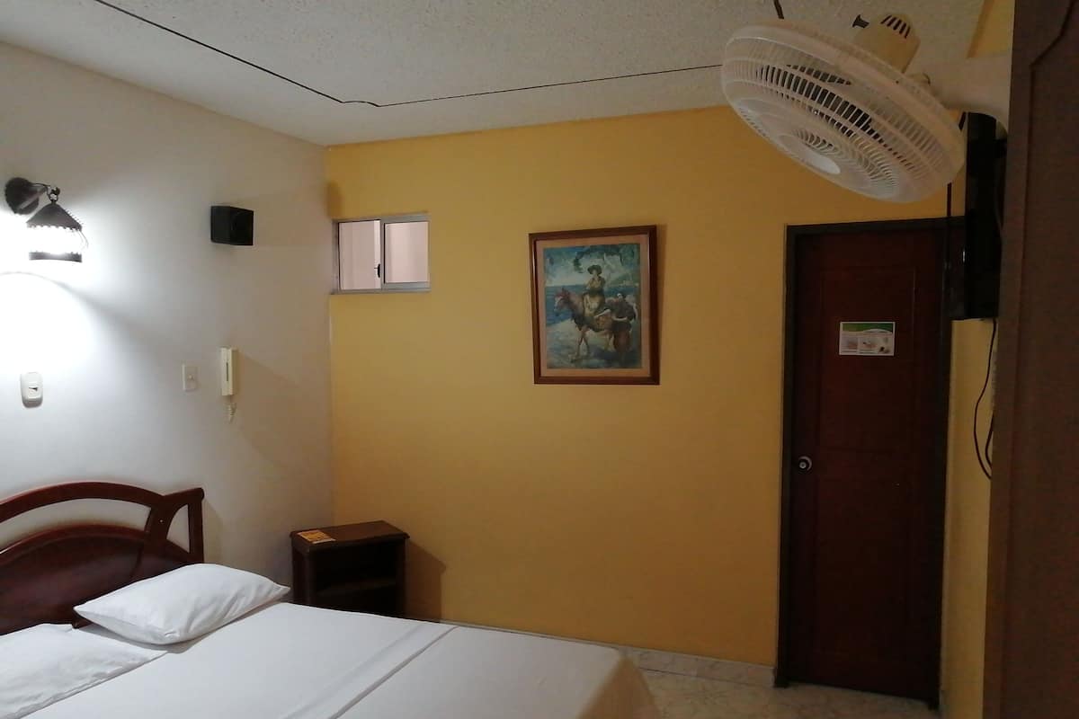 Quarto de hotel com uma cama, uma mesa de cabeceira, um ventilador fixado na parede, uma porta e um quadro.