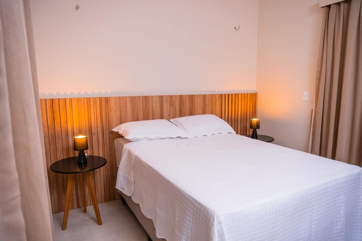 Quarto de hotel com cabeceira de madeira, uma cama de solteiro com roupa de cama branca, duas mesas de cabeceira com luminárias e uma cortina de um lado.