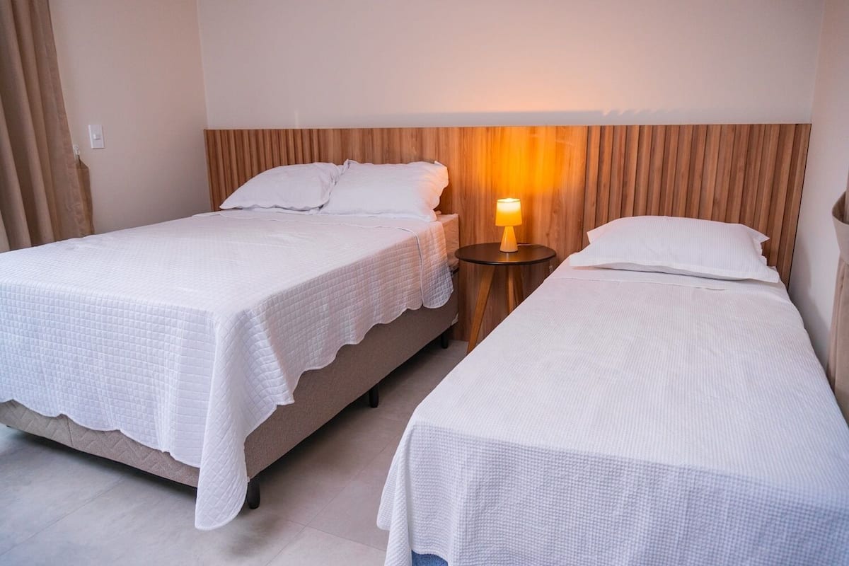 Quarto de hotel com duas camas de solteiro, cabeceira de madeira, mesa de cabeceira com abajur e janela com cortinas.