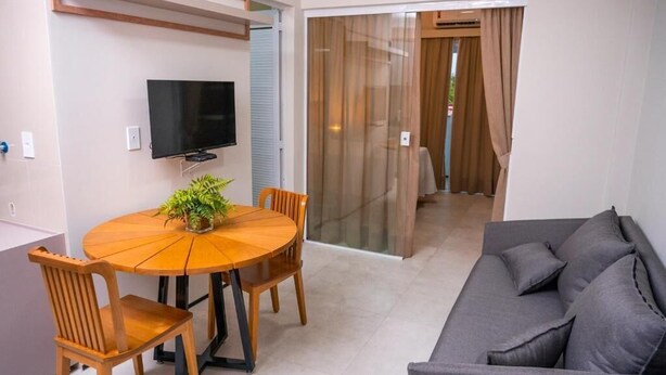 Uma sala de estar moderna com uma mesa de jantar redonda de madeira, quatro cadeiras de madeira, um sofá cinza e uma televisão de tela plana fixada na parede.