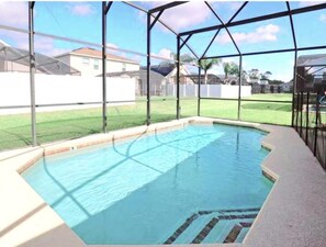 Piscina interior com paredes de vidro transparente, rodeada por uma área coberta com estrutura metálica, e vista para um bairro residencial.