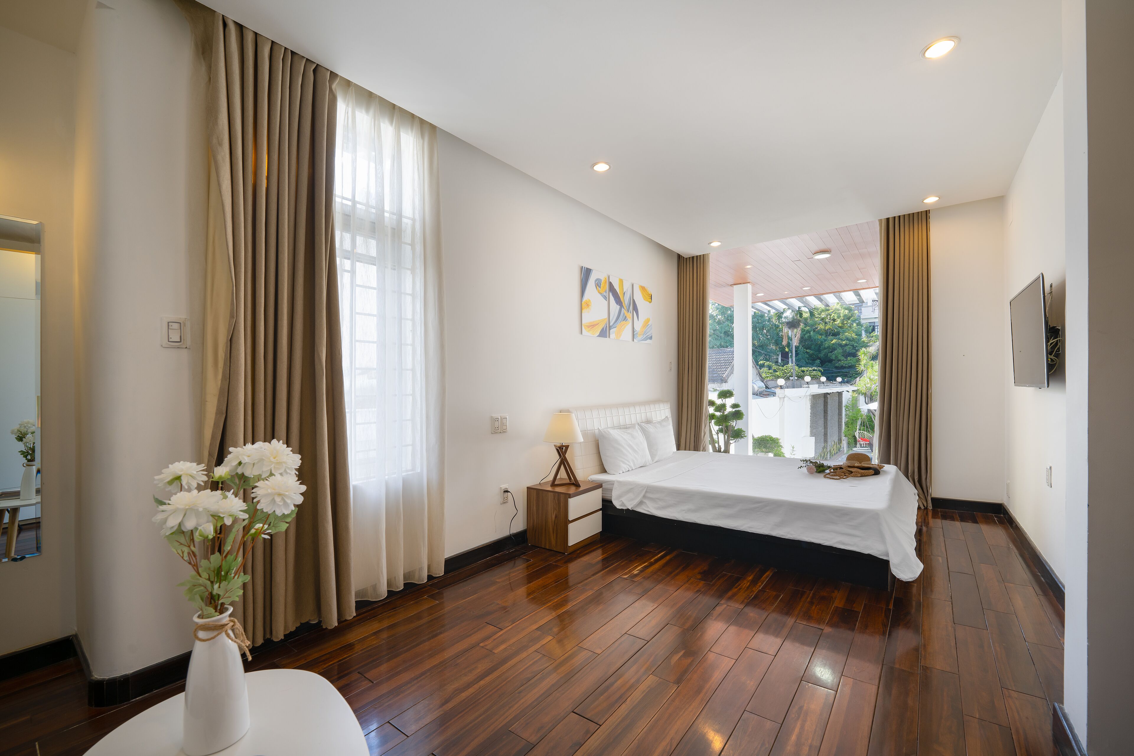 Quarto moderno com uma cama grande, mesa de cabeceira com abajur, vaso com flores e vista para o verde pela janela.
