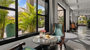 Um hall com uma mesa redonda de mármore, uma poltrona estofada em verde e vista para palmeiras através de grandes janelas.