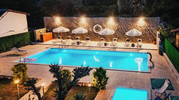 Piscina retangular com espreguiçadeiras e guarda-sóis, cercada por um deck de madeira e vegetação.