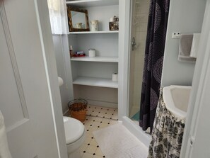 Casa de banho com sanita, lavatório, chuveiro com cortina estampada, prateleira branca com artigos de higiene e chão em azulejos.