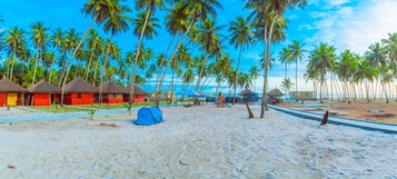 Uma praia com cabanas de palha, uma tenda azul e palmeiras.