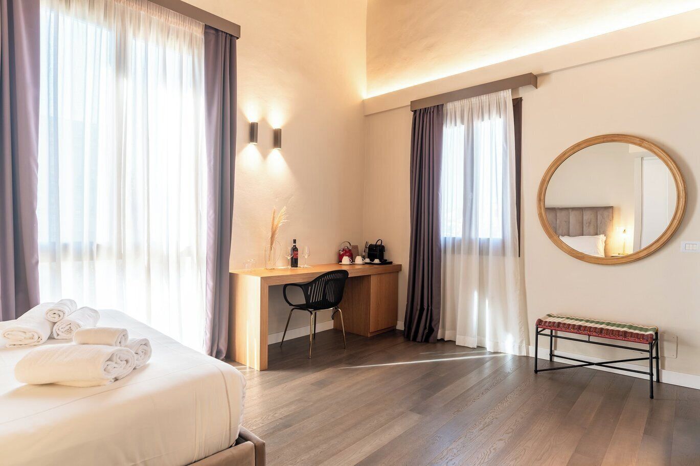Quarto de hotel com uma cama, uma mesa, uma cadeira, um banco e um espelho redondo.