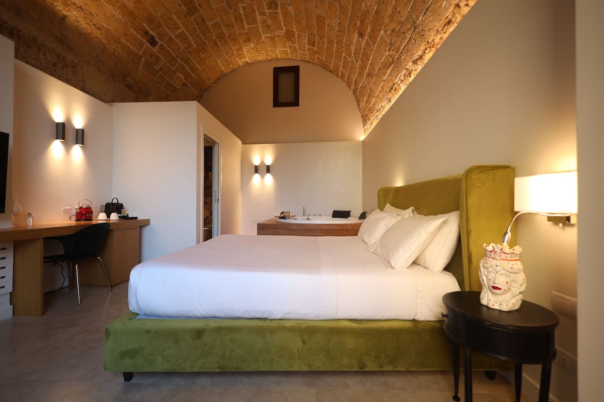Quarto de hotel com teto de tijolos, cama estofada em verde, escrivaninha com cadeira e banheiro com pia e espelho.