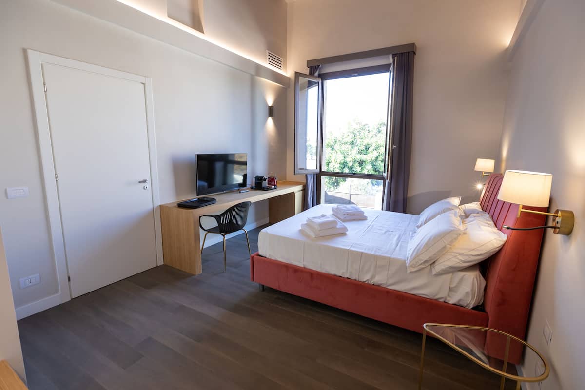 Quarto de hotel com cama, escrivaninha com cadeira, televisão e janela com cortinas.