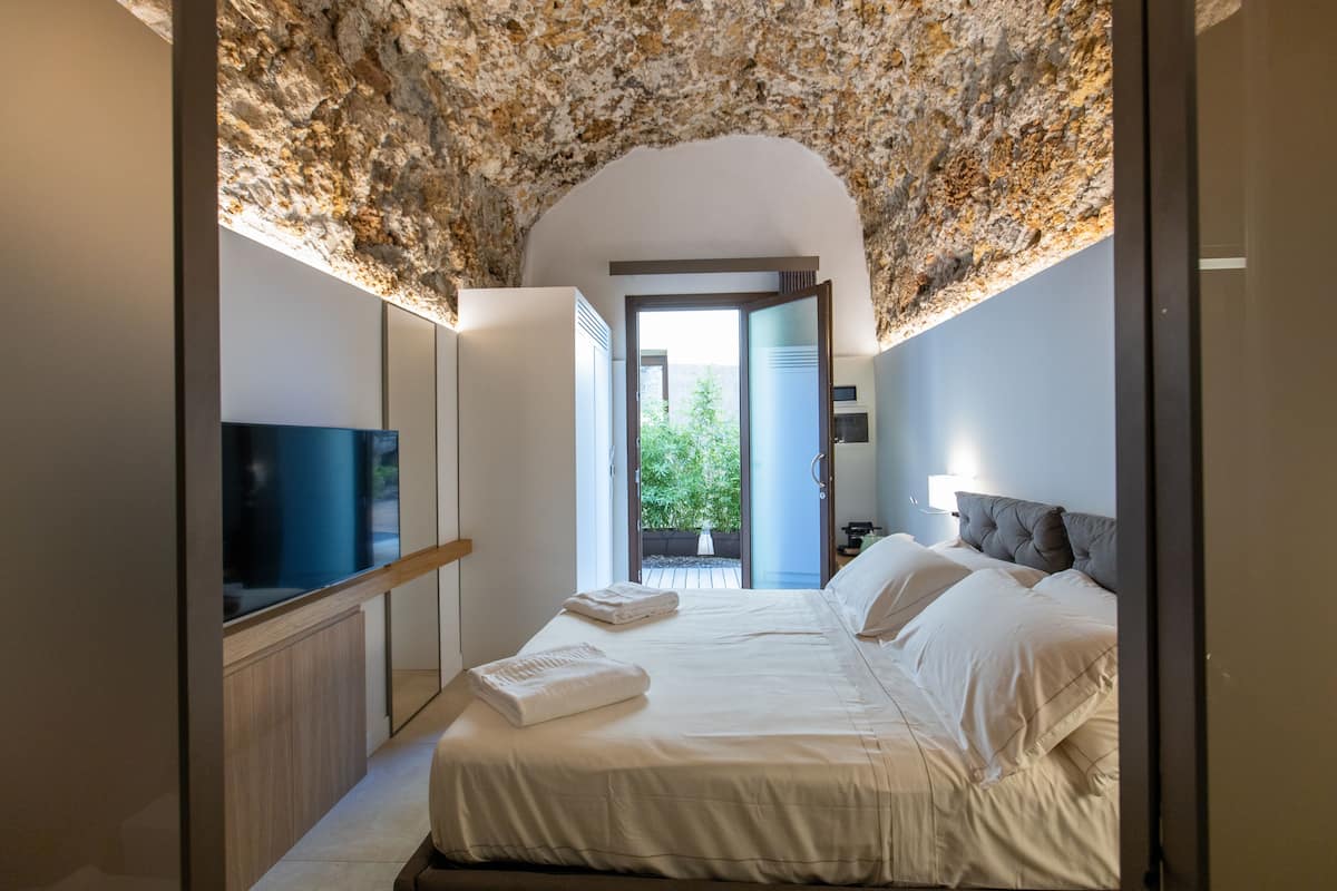 Quarto moderno com teto de pedra, televisão de tela plana, cama com roupa de cama branca e vista para o verde lá fora.