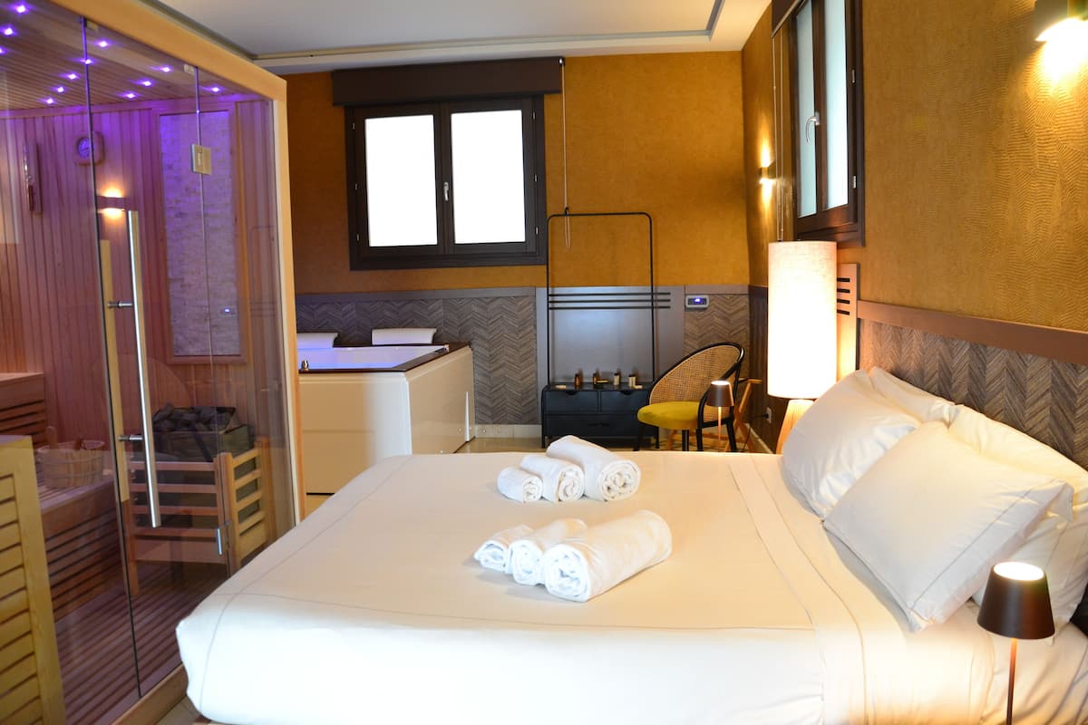 Quarto de hotel com cama, escrivaninha, cadeira e área de duche envidraçada.