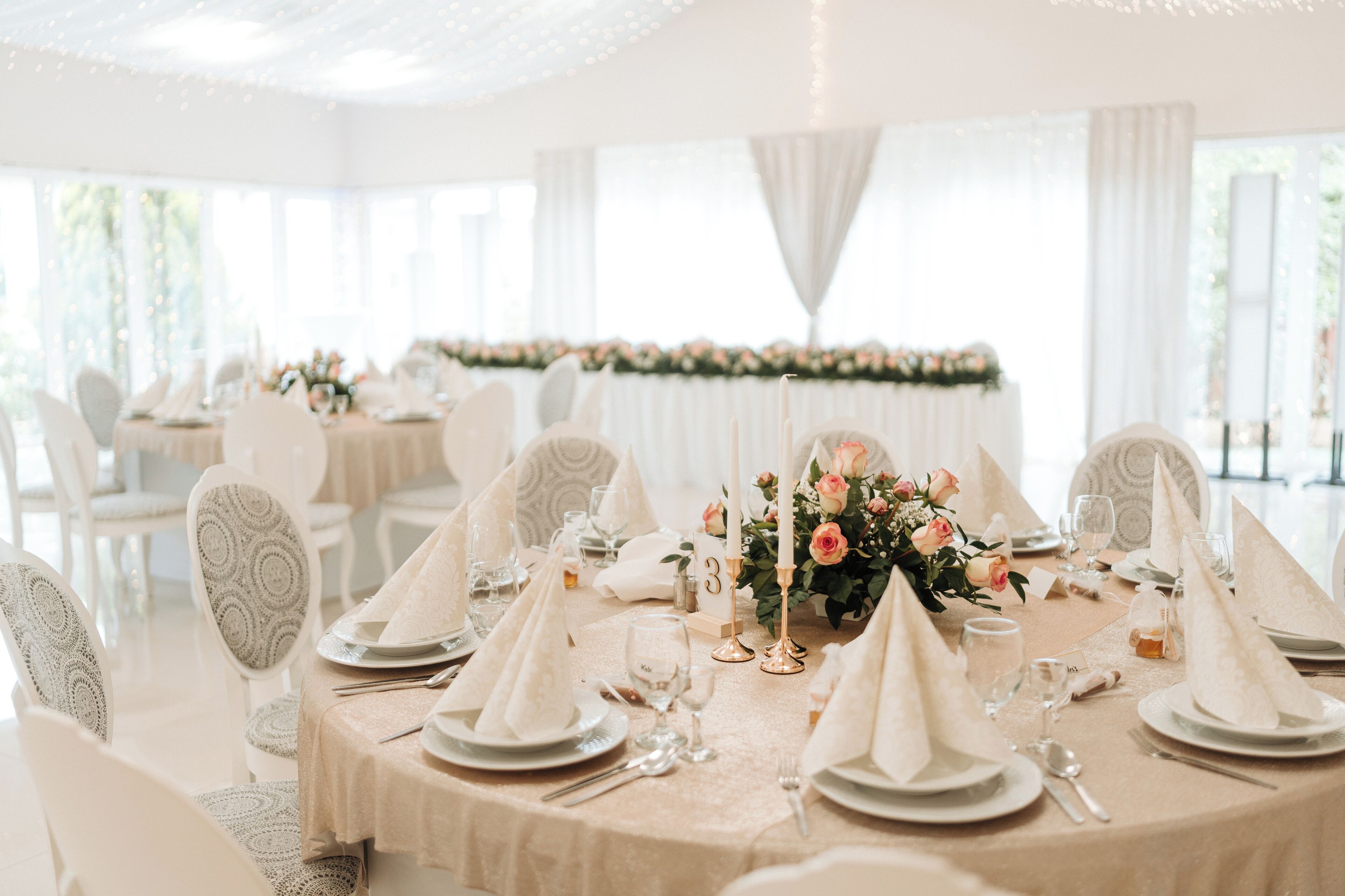 Uma sala de banquetes com mesas redondas dispostas para um evento formal, com toalhas brancas, arranjos florais e louças elegantes.