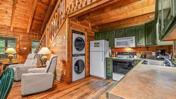 Interior de uma cabana de madeira com cozinha, duas máquinas de lavar roupa, frigorífico e zona de refeições com cadeiras.