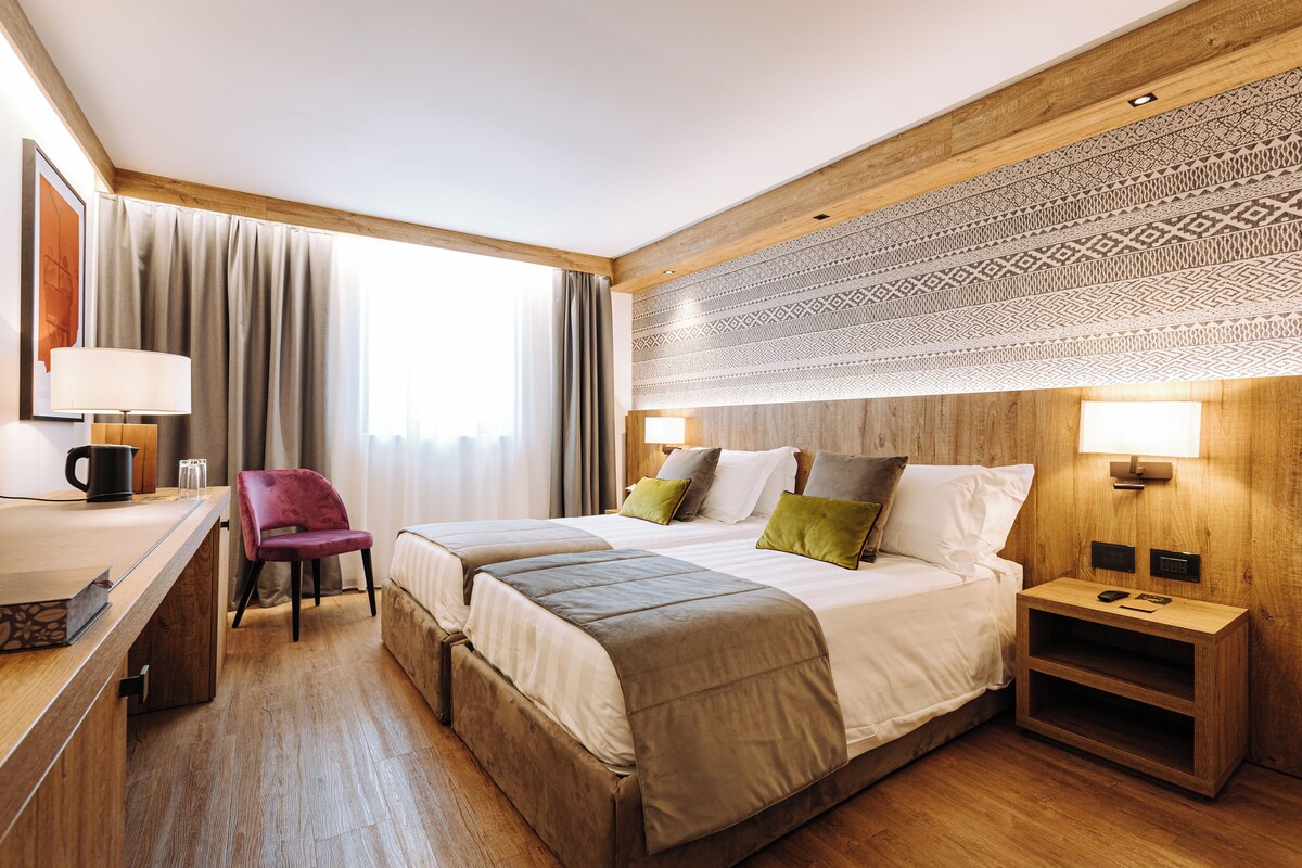 Quarto de hotel com teto de madeira, cama com várias almofadas, escrivaninha com luminária, uma cadeira e mesa de cabeceira com telefone.