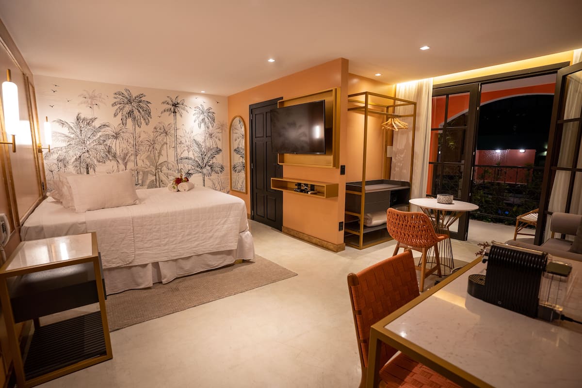 Quarto de hotel com cama, televisão fixada na parede, mesa pequena com cadeiras e vista para o exterior.