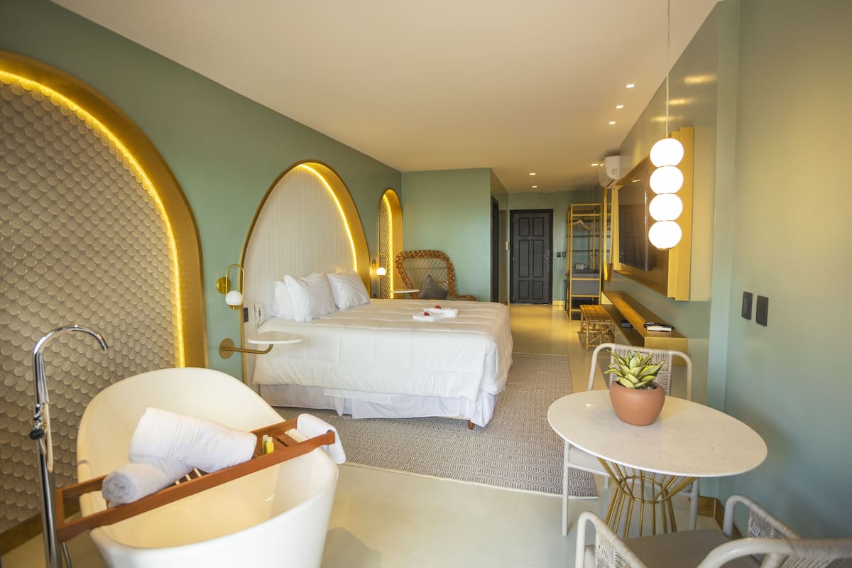Quarto de hotel moderno com uma cama grande, área de estar com uma mesa, banheira e espelho com iluminação.