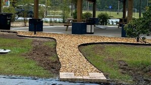 Espaço ao ar livre coberto, com um caminho pavimentado que leva a uma área de estar, cercado por vegetação e um céu claro.