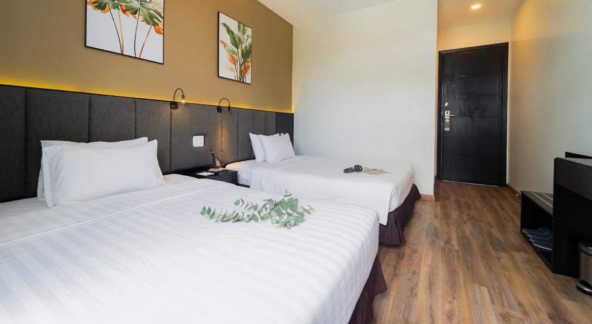 Quarto de hotel com duas camas, cabeceira escura, obra de arte na parede, mesa de cabeceira com abajur e uma porta.