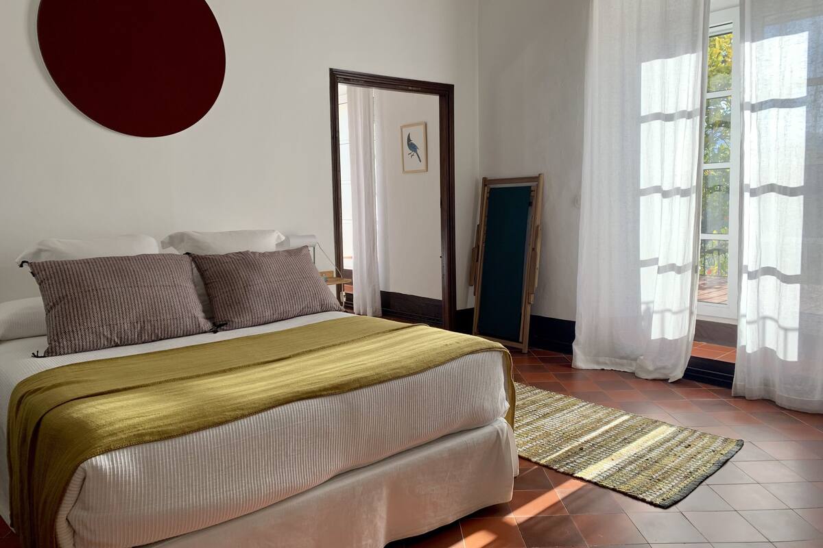 Quarto com cama, almofadas, um grande ornamento circular na parede, uma porta com espelho e vista para o exterior através de cortinas translúcidas.